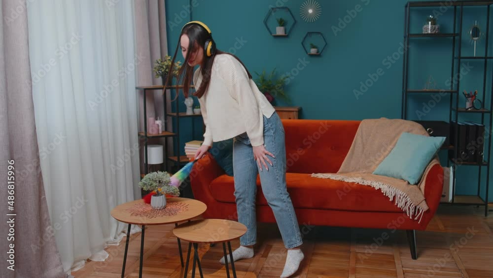 Joyful young woman dusting furniture caring for hygiene using colorful duster in living room ...