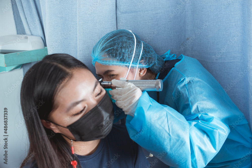 A doctor in full PPE gear checks the ear drum of a female patient with ...