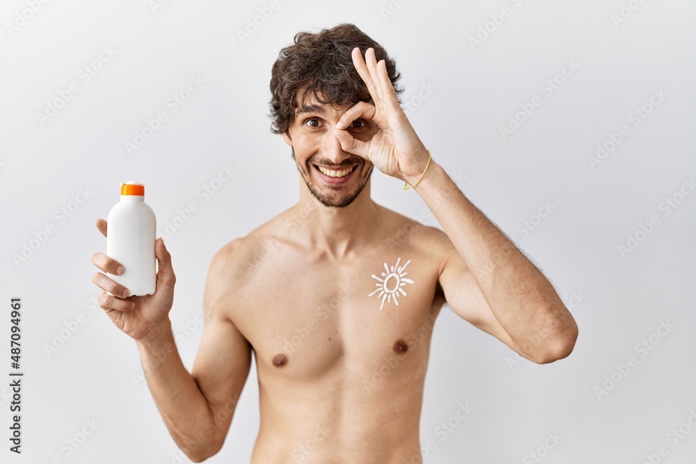 Young hispanic man standing shirtless holding sunscreen lotion doing ok ...