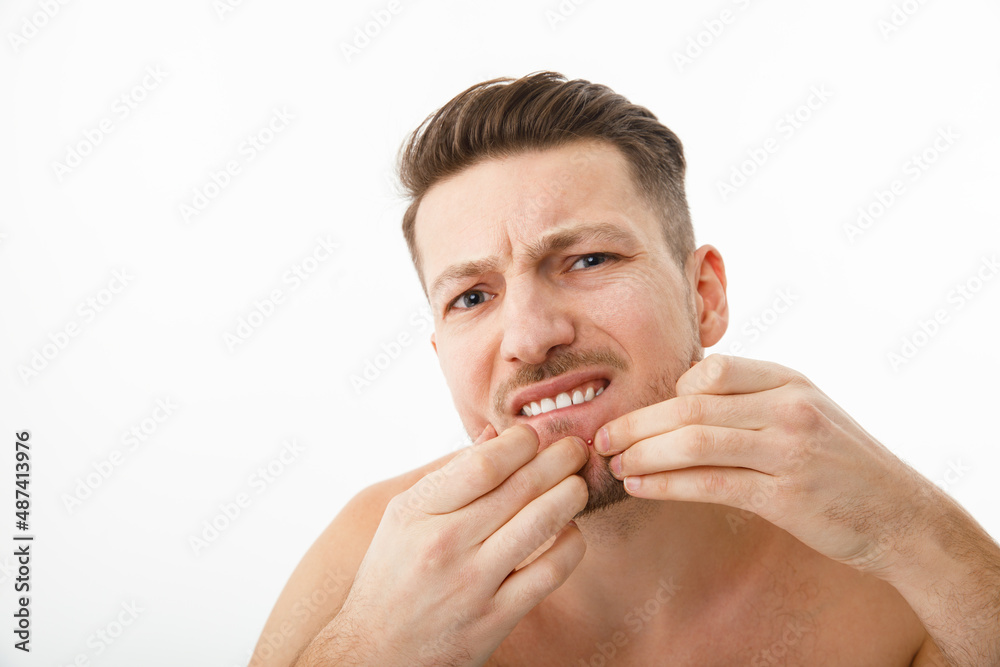 A young man squeezes out a pimple on his face while looking in the ...