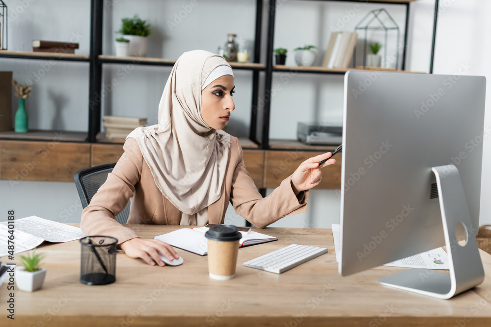 muslim woman pointing at computer monitor while working in home office ...