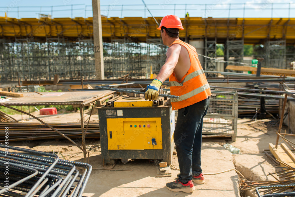 Builder in a hard hat and vest bending rebar on a bending machine at a ...