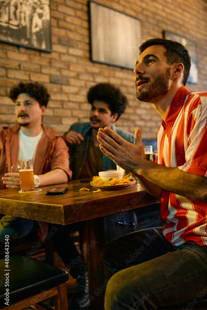 Multiracial group of young sports fans watching match on TV in a pub ...