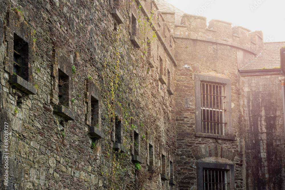 Old stone jail wall with small windows with metal bars. Sun flare. Law correction facility with ...