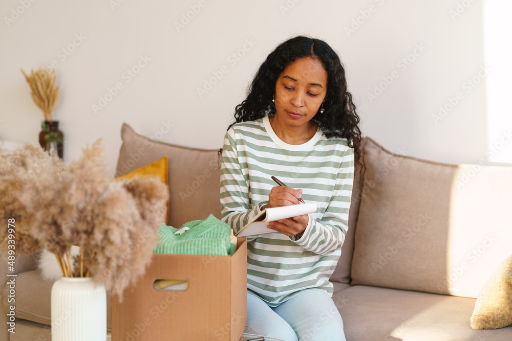 African-american female keeping count for clothing in cardboard box. Moving away and packing ...