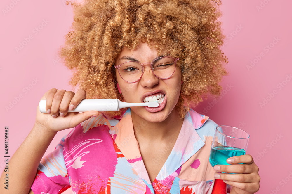 Horizontal shot of young woman with curly hair brushes teeth with ...