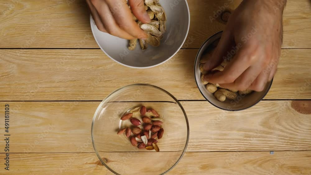 Viewed from above, men's hands peel peanuts from their shells, tossing ...