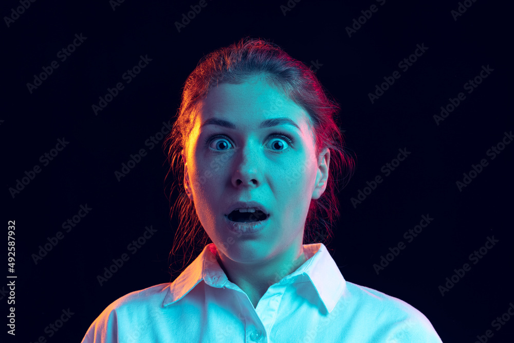 Shocked young girl in white shirt looking at camera with open mouth isolated on dark background ...
