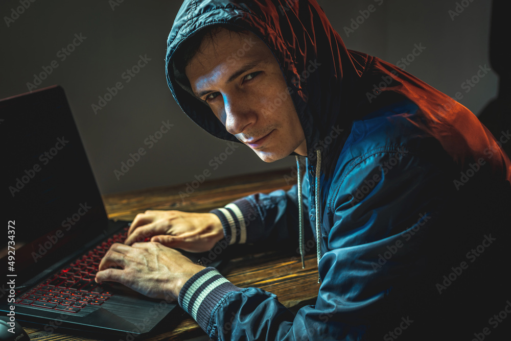 Hacker in a hood with a phone is typing on a laptop keyboard in a dark room. Concept of cyber ...