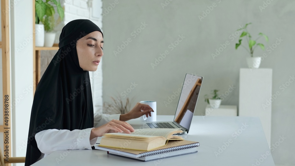 A Muslim female teacher reads a book and types learning material on a ...
