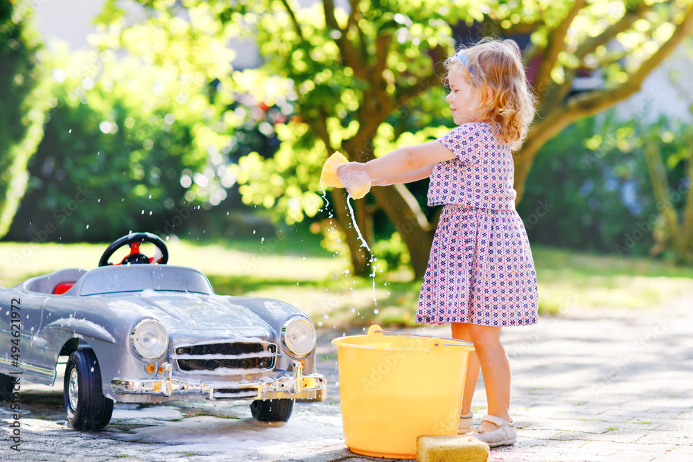 Cute gorgeous toddler girl washing big old toy car in summer garden ...