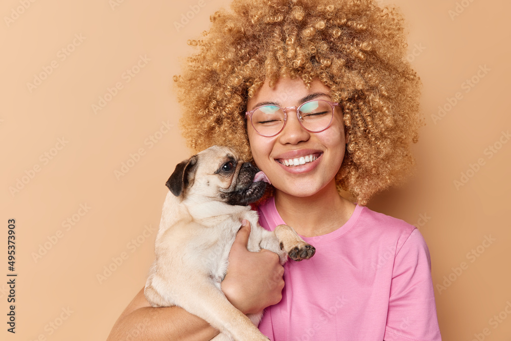 Happy curly haired woman receives kiss from favorite pet smiles broadly ...