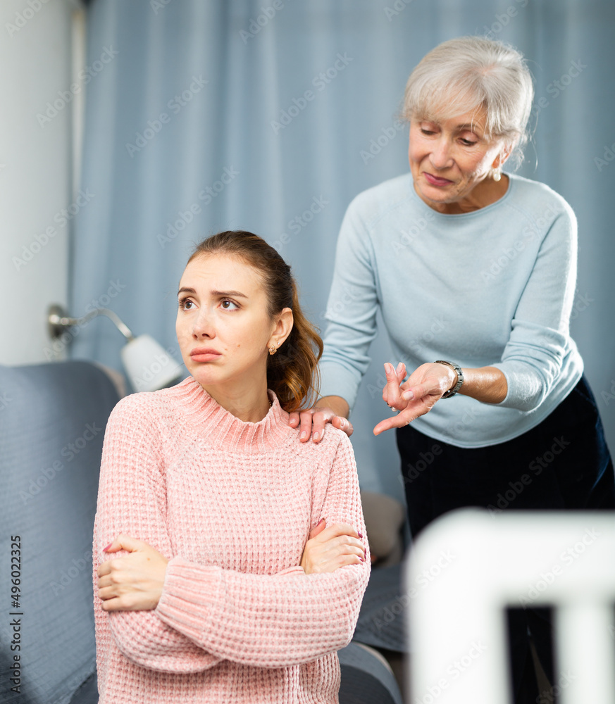 Frustrated young girl sitting on sofa at home while agitated elderly mother reprimanding her ...