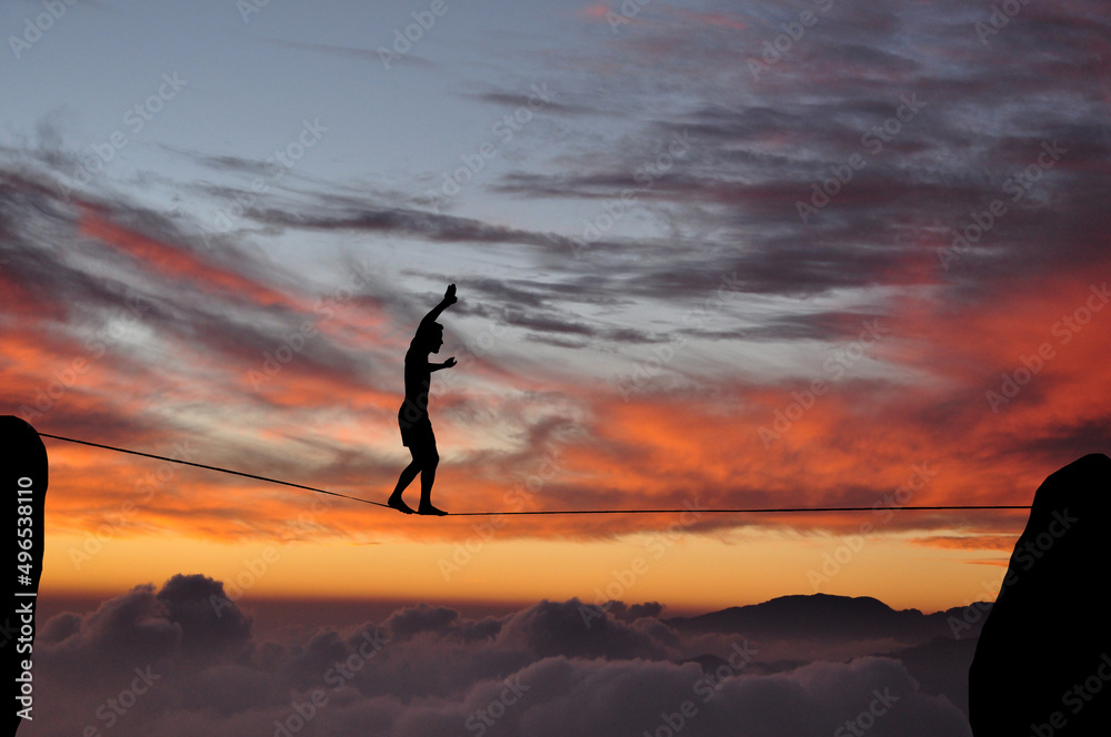 Silhouette of young man balancing on slackline high above clouds and ...