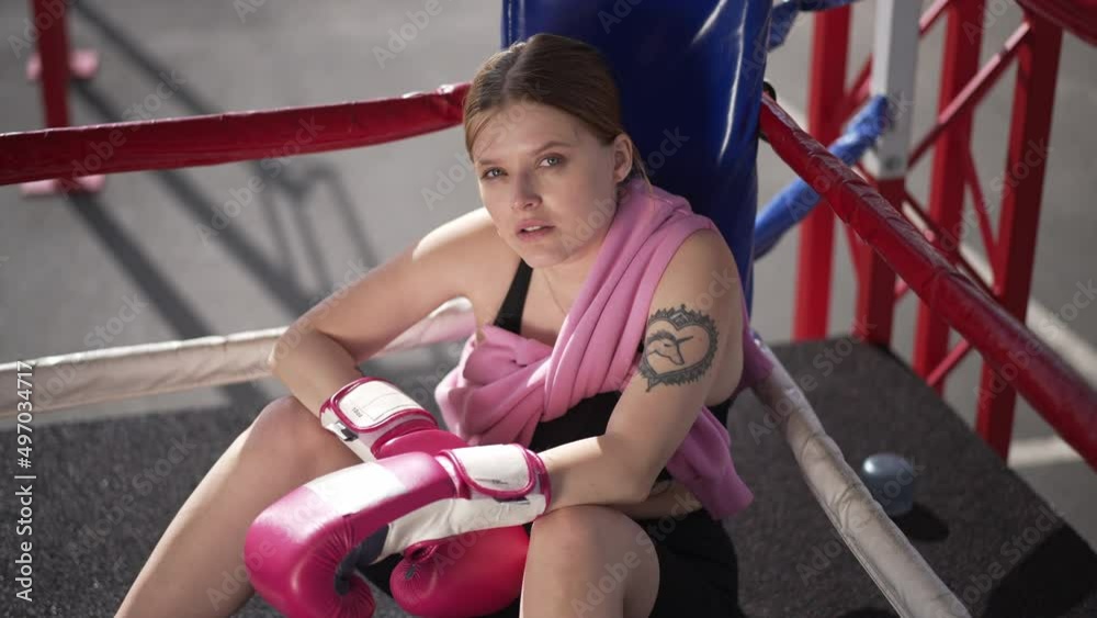 Exhausted fit sportswoman in boxing gloves sitting on ring looking at ...