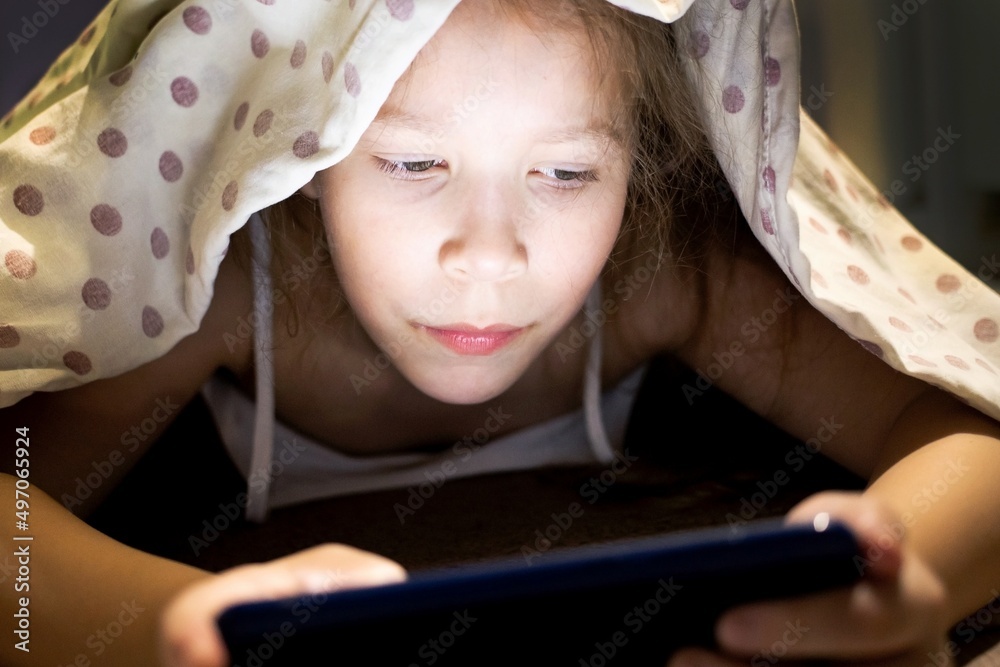 Foto de Cute little girl hiding under a blanket and watching a cartoon ...