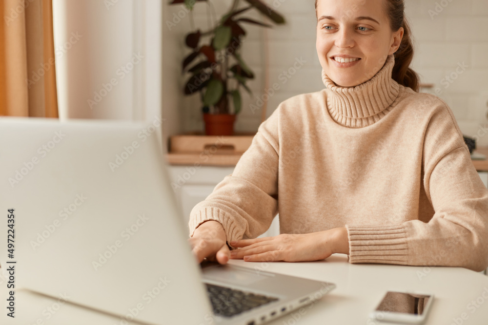 Happy pretty woman wearing beige jumper posing in kitchen and working ...
