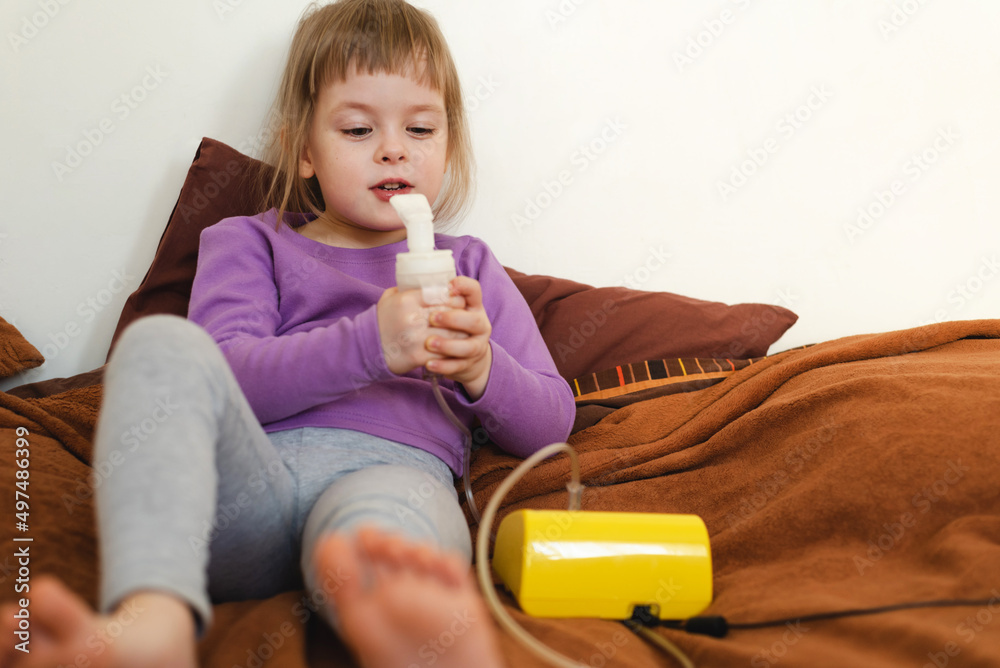 Sick child lies in bed and makes inhalation using a nebulizer. The girl ...