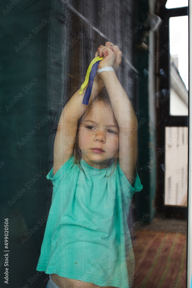 a sad little Ukrainian girl is standing behind the glass with her hands ...