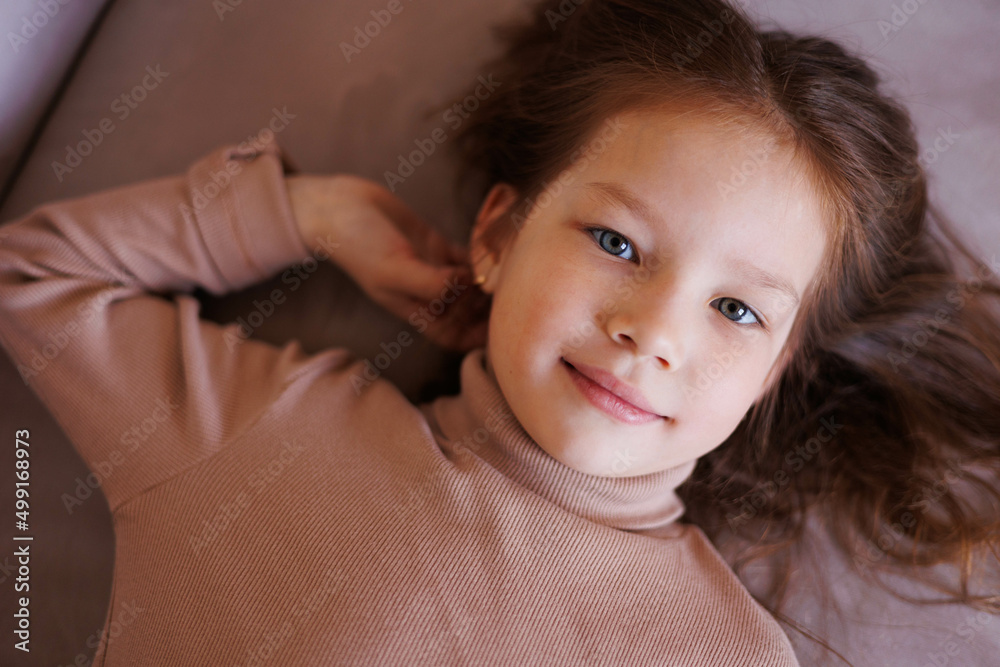 Portrait of 6-year-old little girl laying on her back in casual outfit, on sofa at home. Female ...