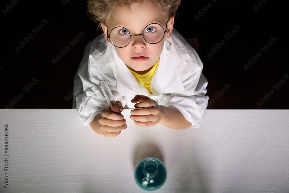 Serious little boy in white uniform and glasses conducts chemical ...