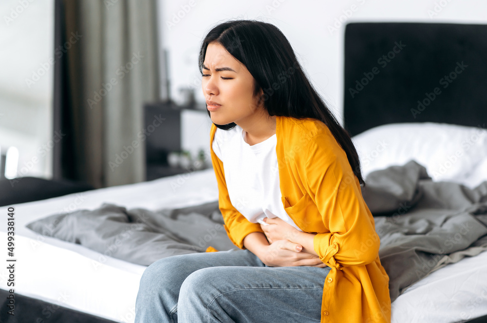 Asian sad brunette girl, sitting at home on the bed, holding her hands ...