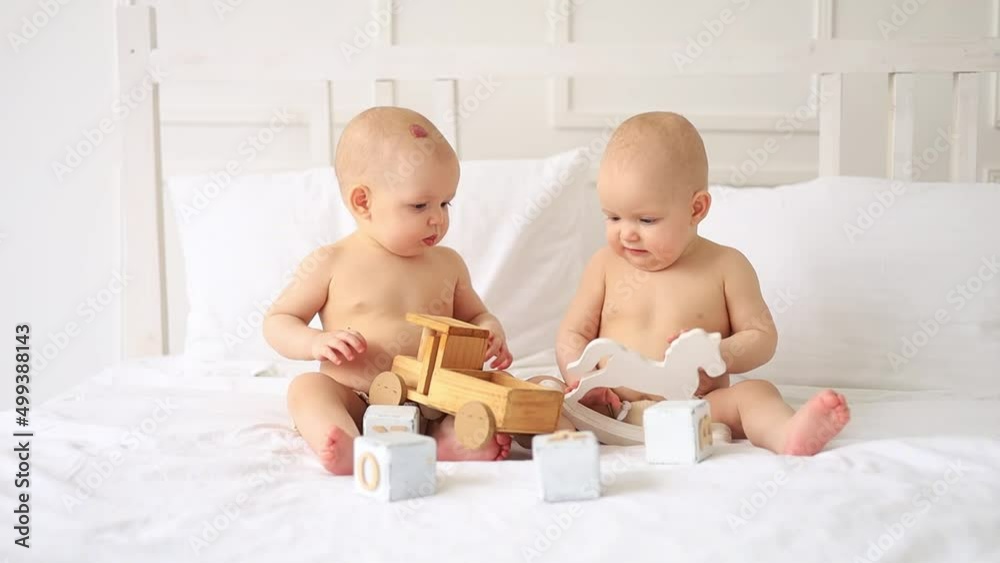 two twin babies in diapers play with wooden toys at home on a bed in a ...
