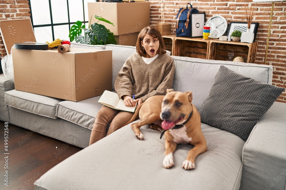Young beautiful woman sitting on the sofa at new home scared and amazed ...