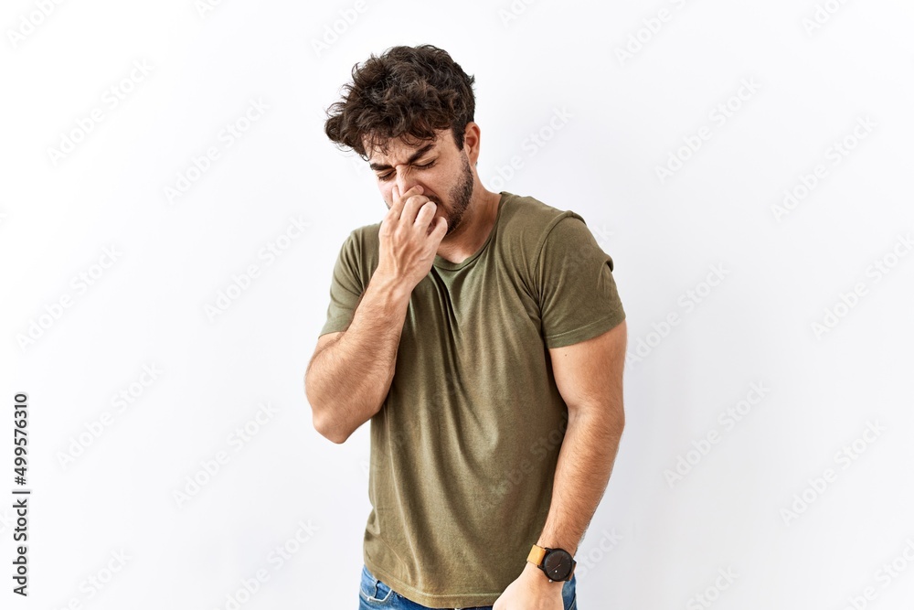 Hispanic man standing over isolated white background smelling something ...