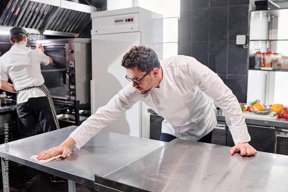Chef in uniform wiping kitchen table after cooking with his colleague ...