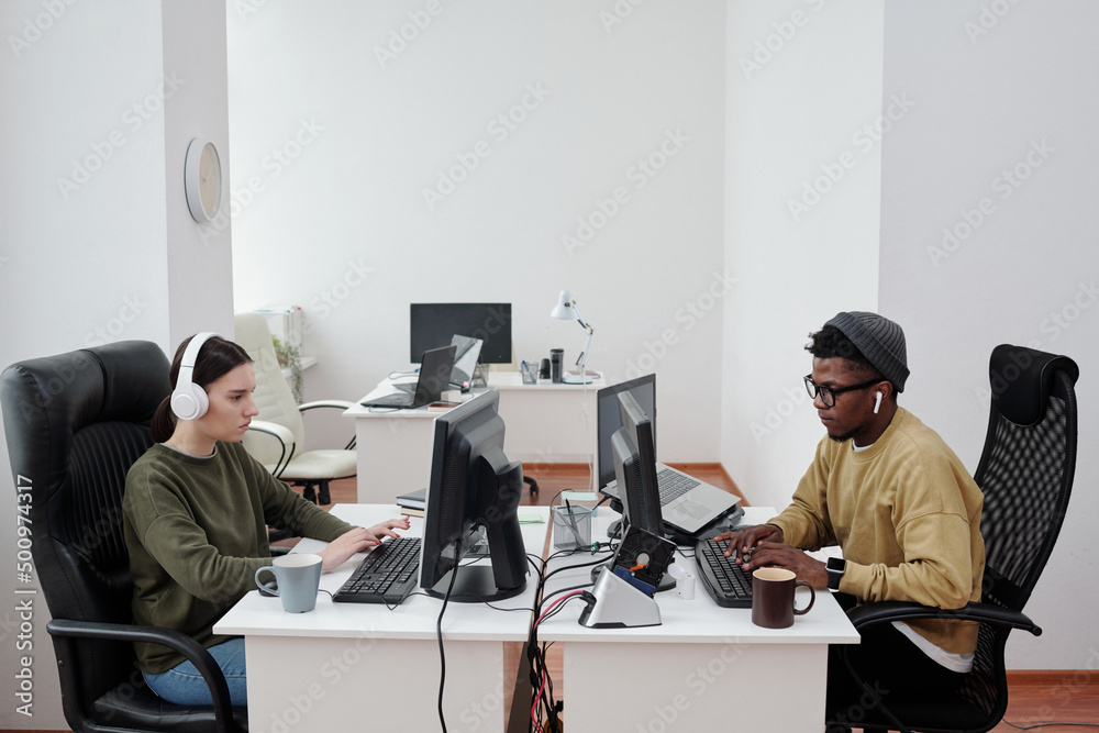Side view of two young intercultural programmers working in front of computers while decoding ...