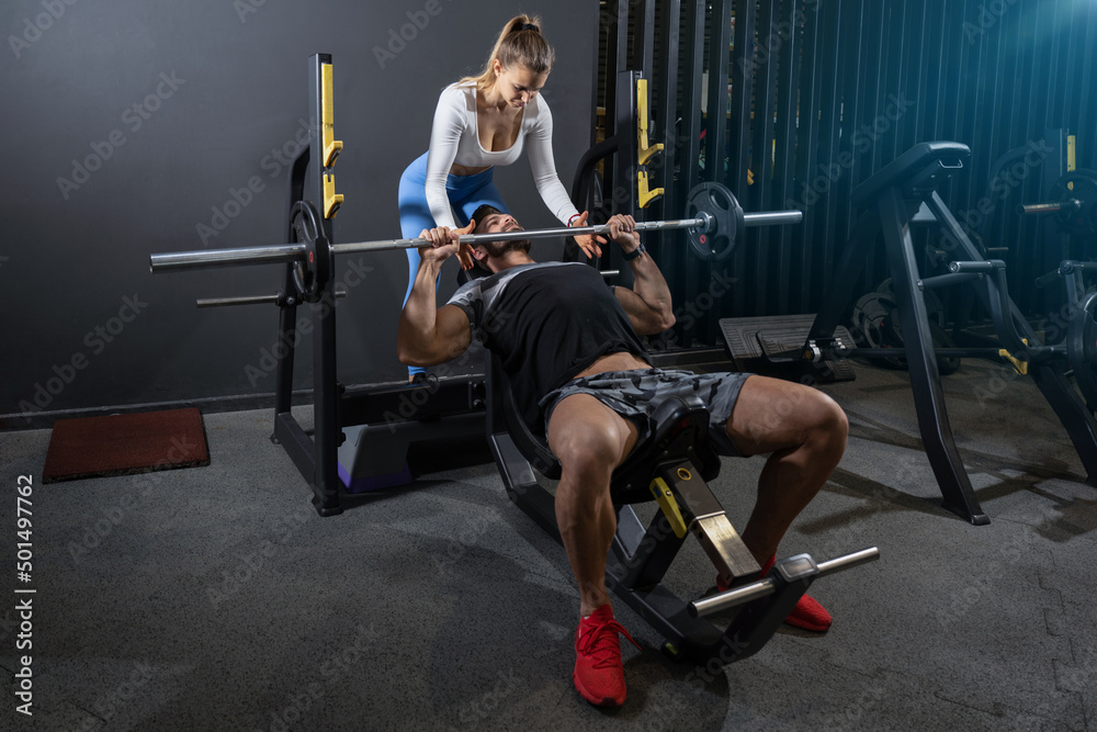 Muscular young man lifting barbells with assistance of attractive young ...