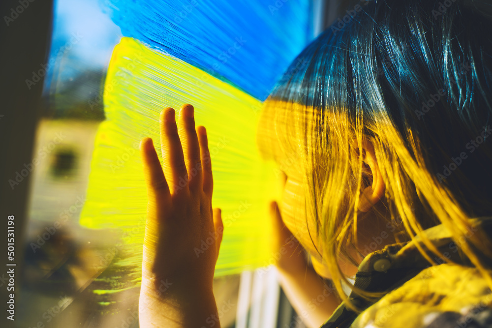 Little child with hands on yellow-blue flag image of Ukraine on window ...