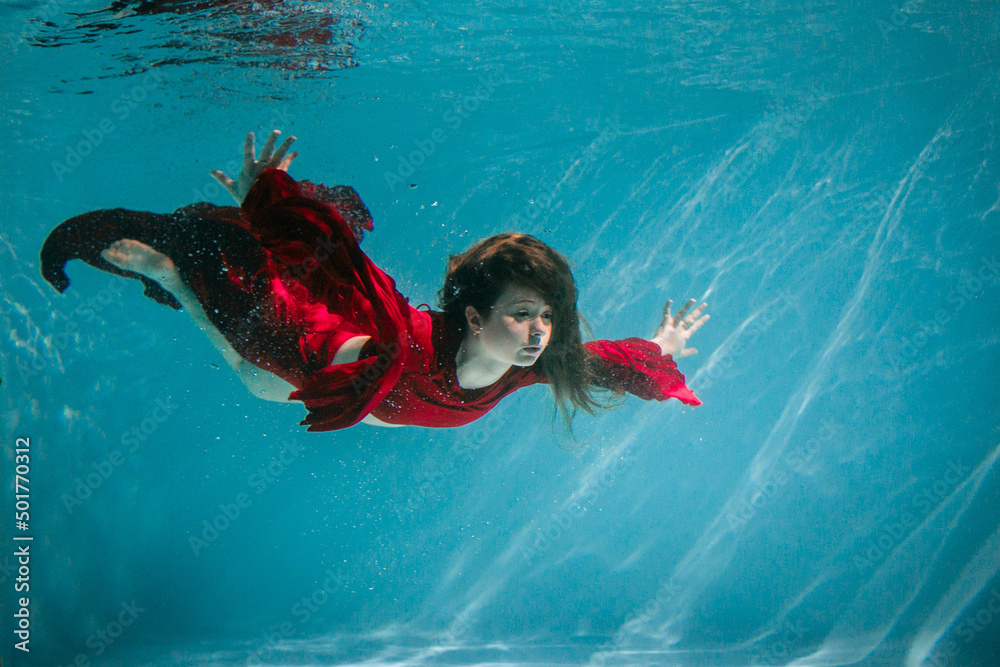Attractive red-haired young woman swims beautifully underwater in a red ...