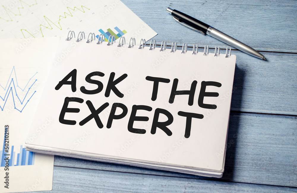 Text ASK THE EXPERT on the page of a notepad lying on financial charts ...