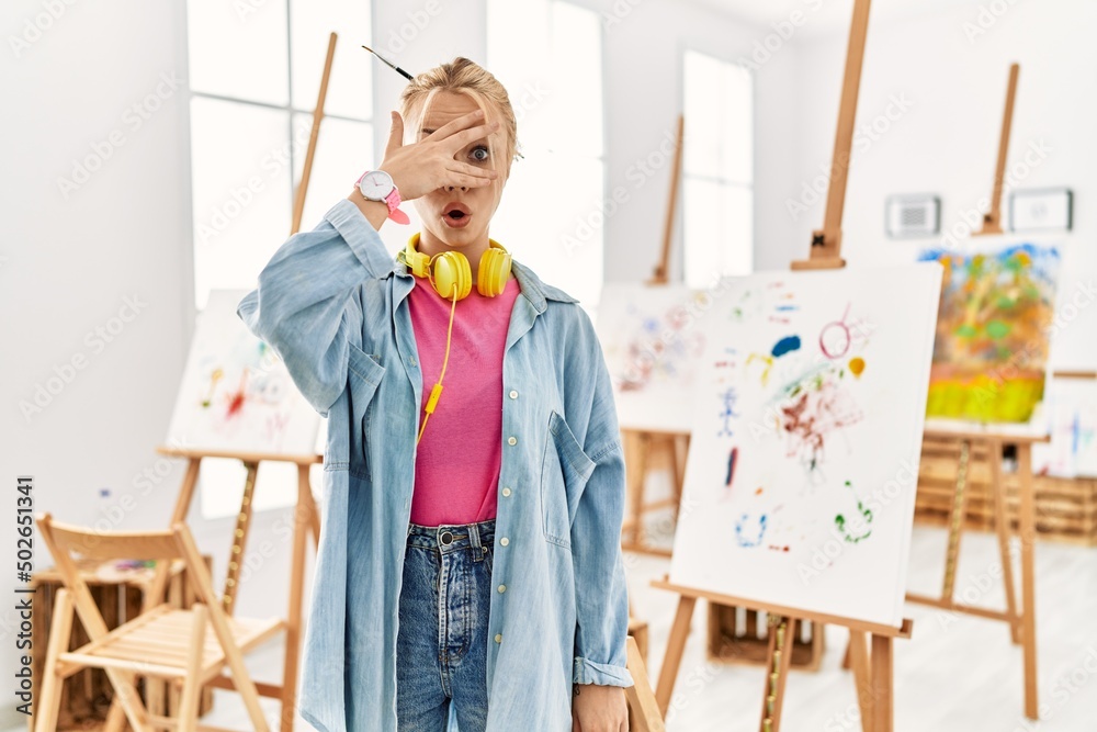 Young caucasian girl at art studio peeking in shock covering face and ...