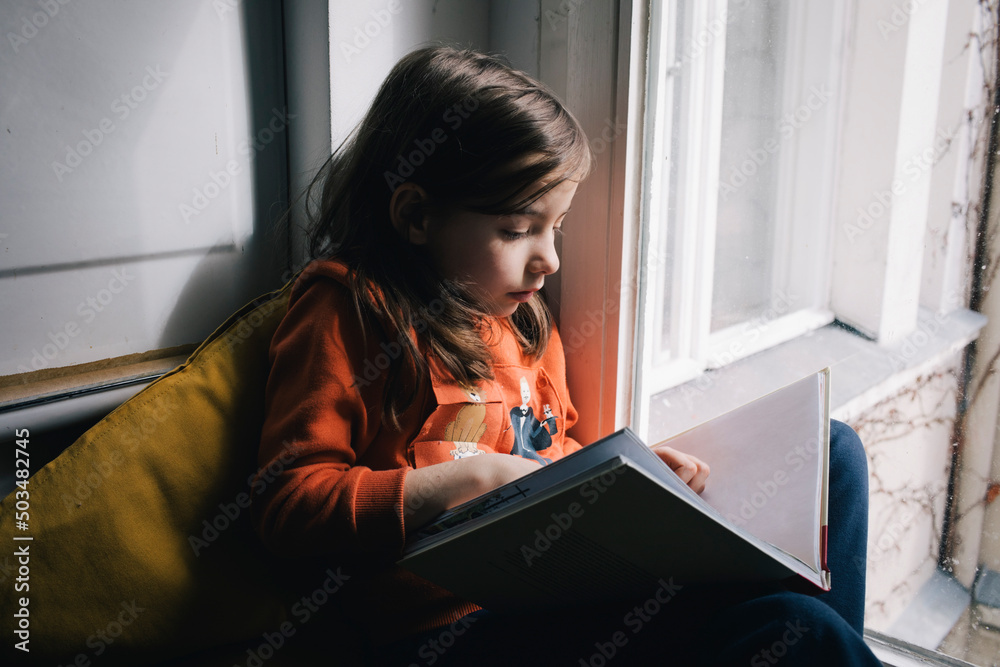 Girl reading book sitting by glass window at home Stock Photo | Adobe Stock
