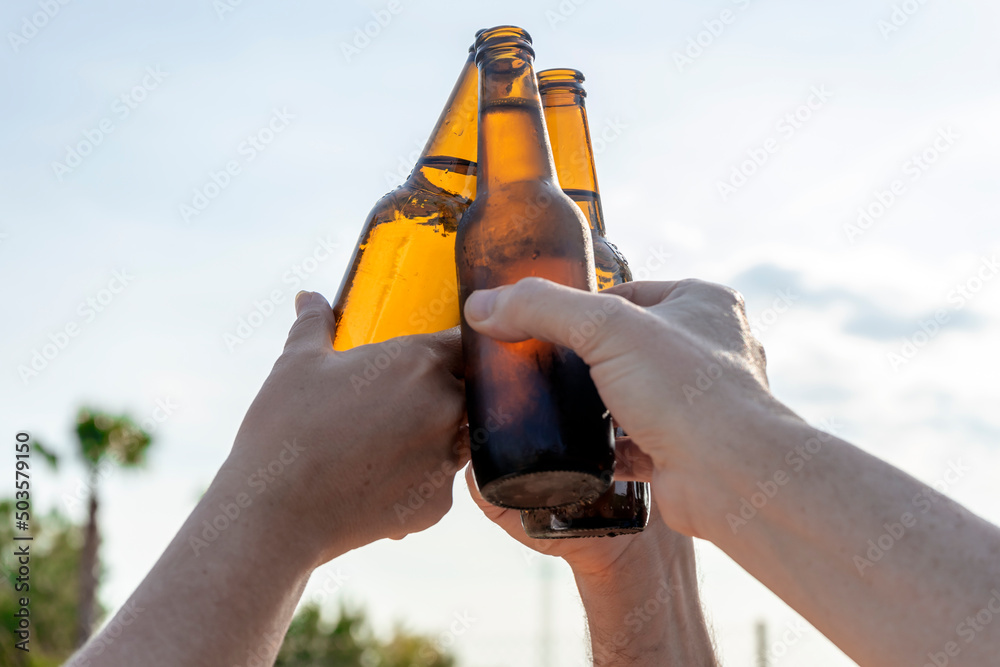 FRIENDS CELEBRATING AND CLINKING BEER BOTTLES OUTDOOR AT SUNSET ...
