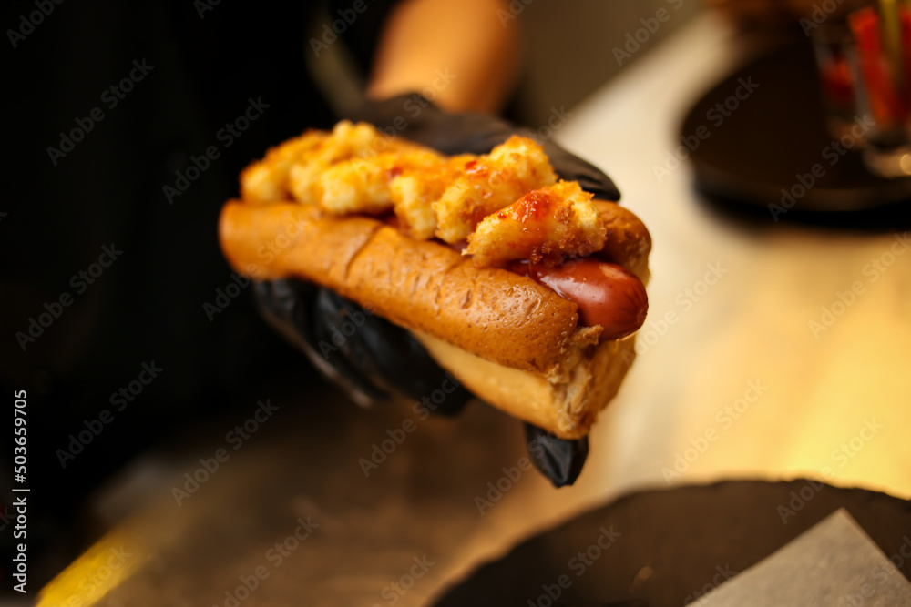 cook in black gloves holds hot dog. The cook prepares a hot dog, adding ...