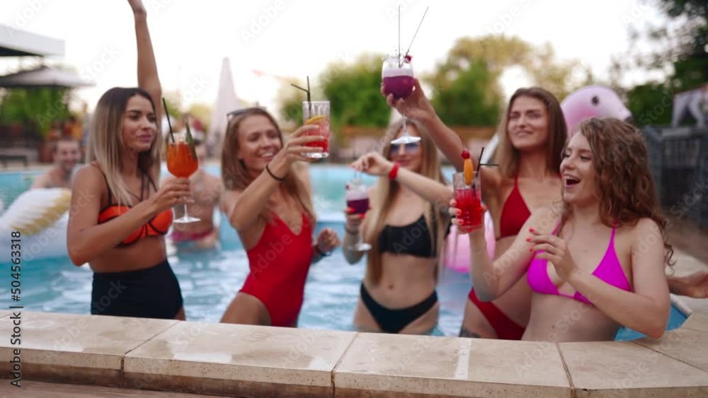 Girls in bikini have pool party with cocktails in swimming pool. Women relaxing clinking glasses ...
