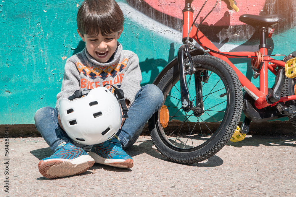 happy boy with bycicle and helmet sitted on the ground of the city ...