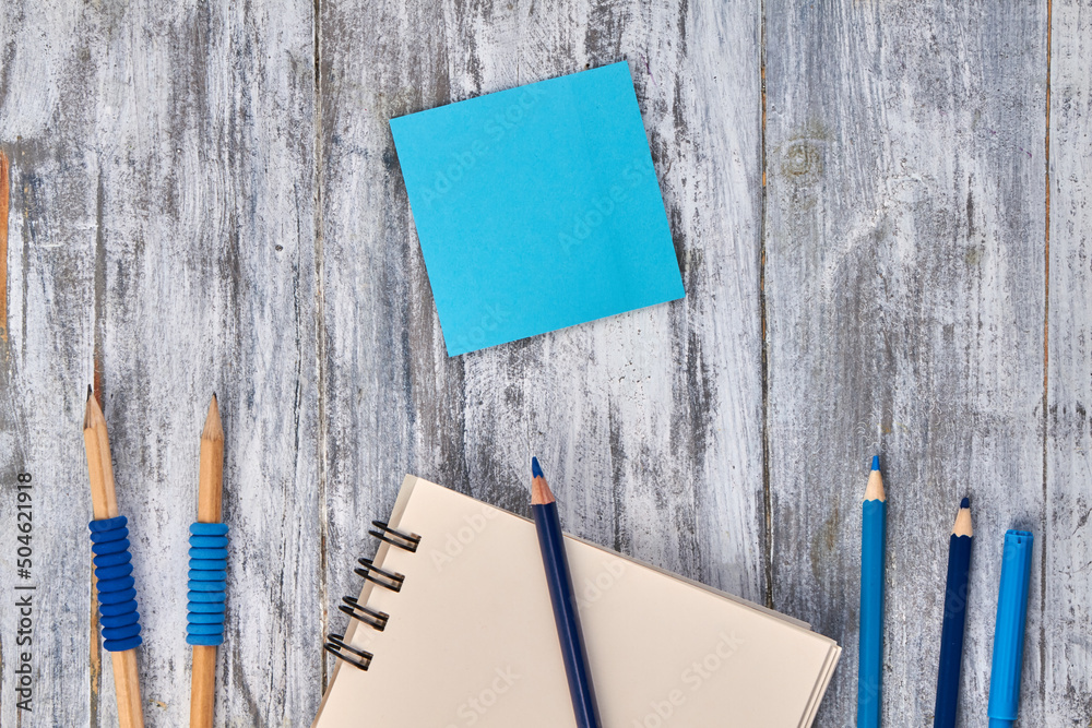 Blank blue sticky note with pencils and notepad. Gray wood background ...