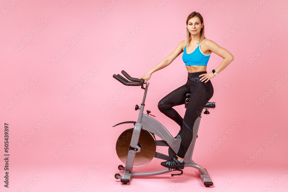 Beautiful blonde woman sitting on exercise bike, looking at camera ...