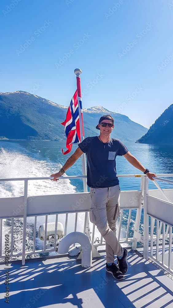 A young man leaning on a rail of a ship with a Norwegian flag waving ...