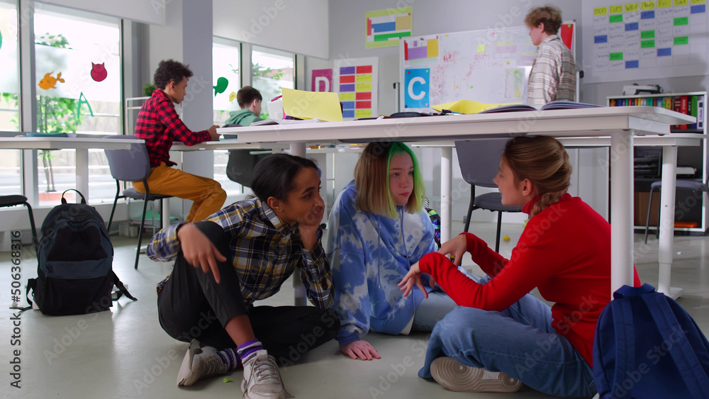 Happy Diverse Teen Girlfriends Sit Under Desk In Class And Gossip During Break Stock Foto