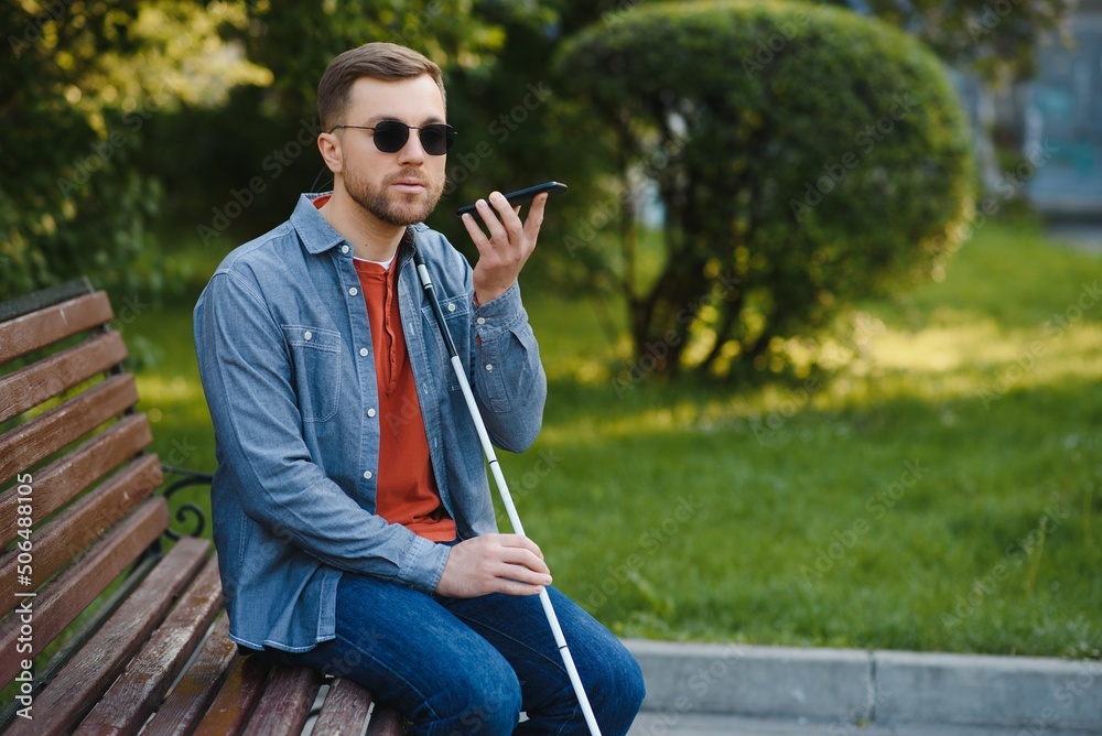 Young blind man with smartphone sitting on bench in park in city ...