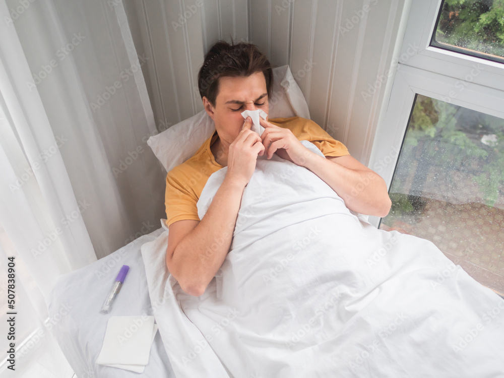 Sick man is lying in bed with thermometer. Man is blowing his nose ...