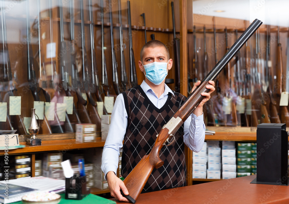 Seller in medical face mask standing behind counter at gun shop ...
