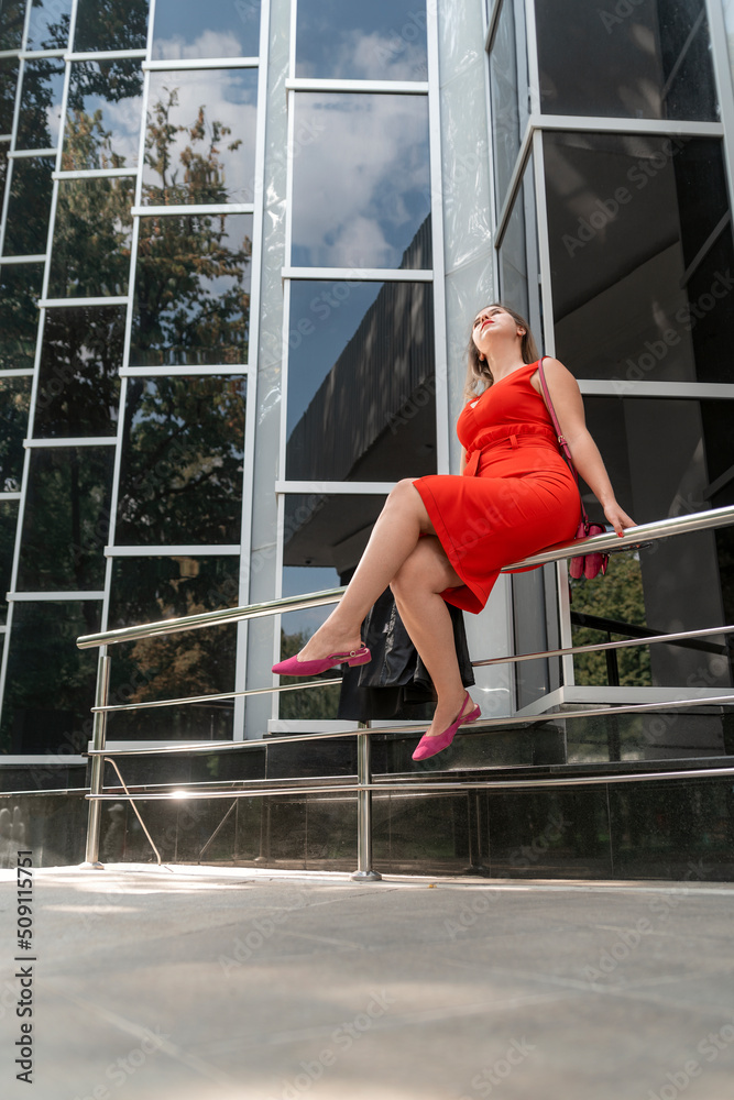 Portrait of plump young woman in red dress sits on railing on modern ...