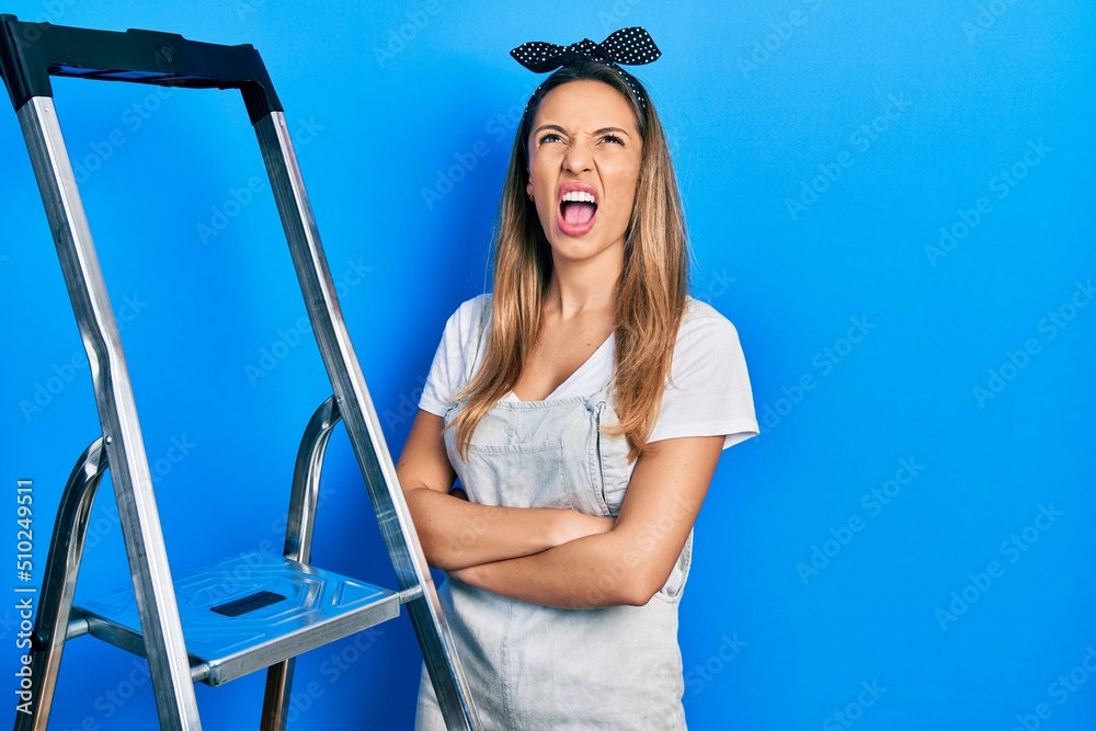 Beautiful hispanic woman standing by ladder with crossed arms angry and ...